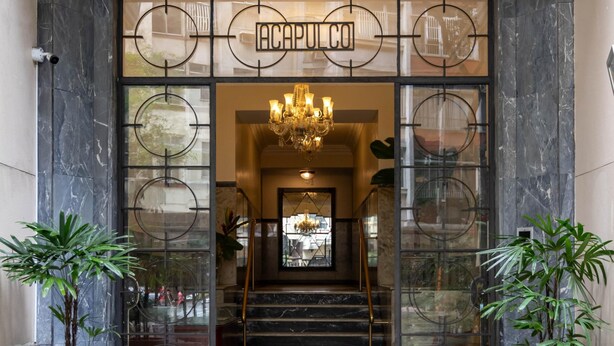 Entrada de hotel com fachada de mármore, um lustre e plantas em vasos.