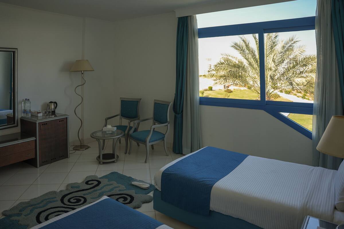 Quarto de hotel com duas camas, uma escrivaninha, uma cadeira, uma mesa pequena, um abajur e vista para palmeiras.