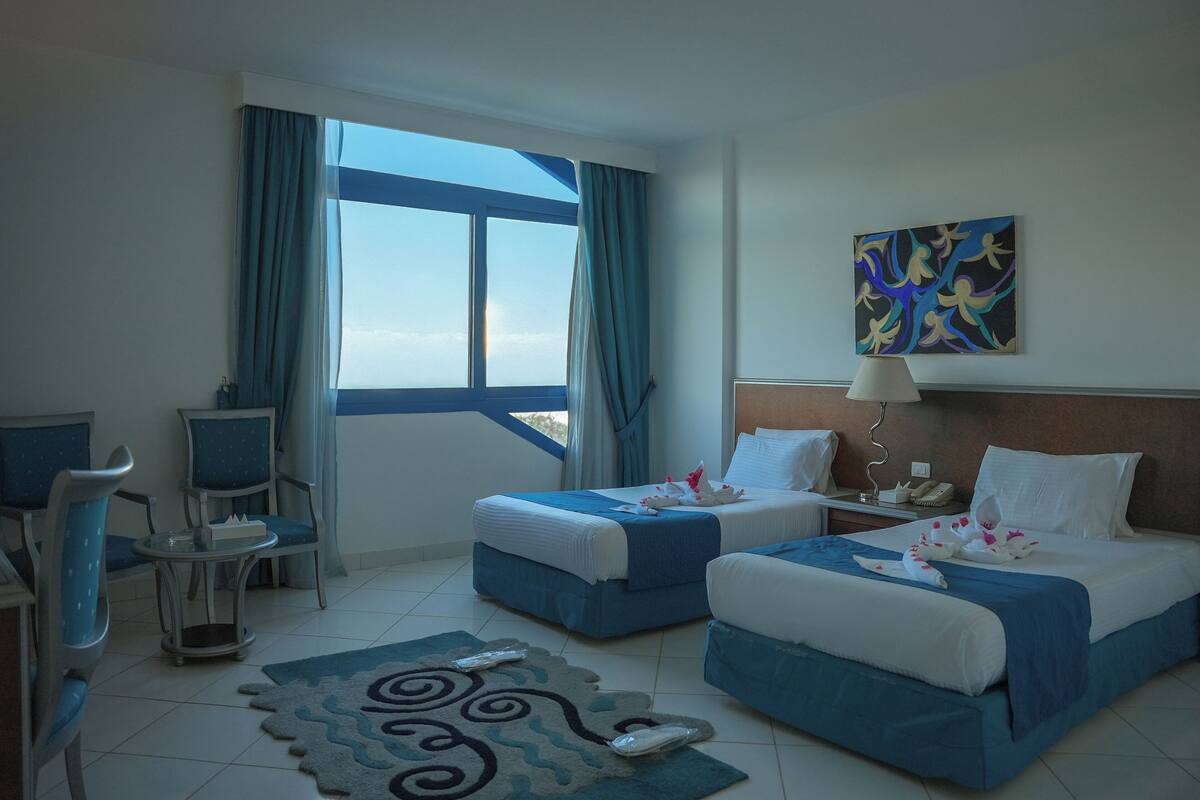 Quarto de hotel com duas camas, uma escrivaninha, uma cadeira, uma janela com cortinas e um quadro na parede.