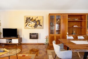 Uma sala de estar moderna com televisão de tela plana, um armário de madeira com portas de vidro, uma mesa de jantar com cadeiras e um grande quadro abstrato na parede.