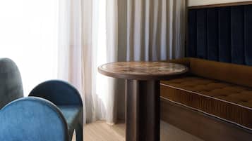 Quarto de hotel moderno com mesa redonda, duas cadeiras azuis, um sofá e uma janela grande com cortinas leves.