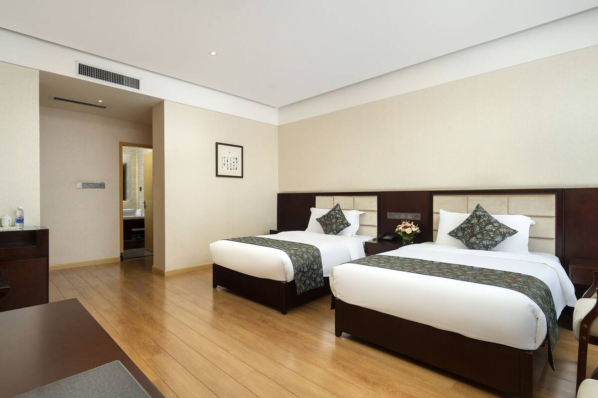 Quarto de hotel com duas camas, uma escrivaninha, televisão e banheiro visível através de uma porta aberta.