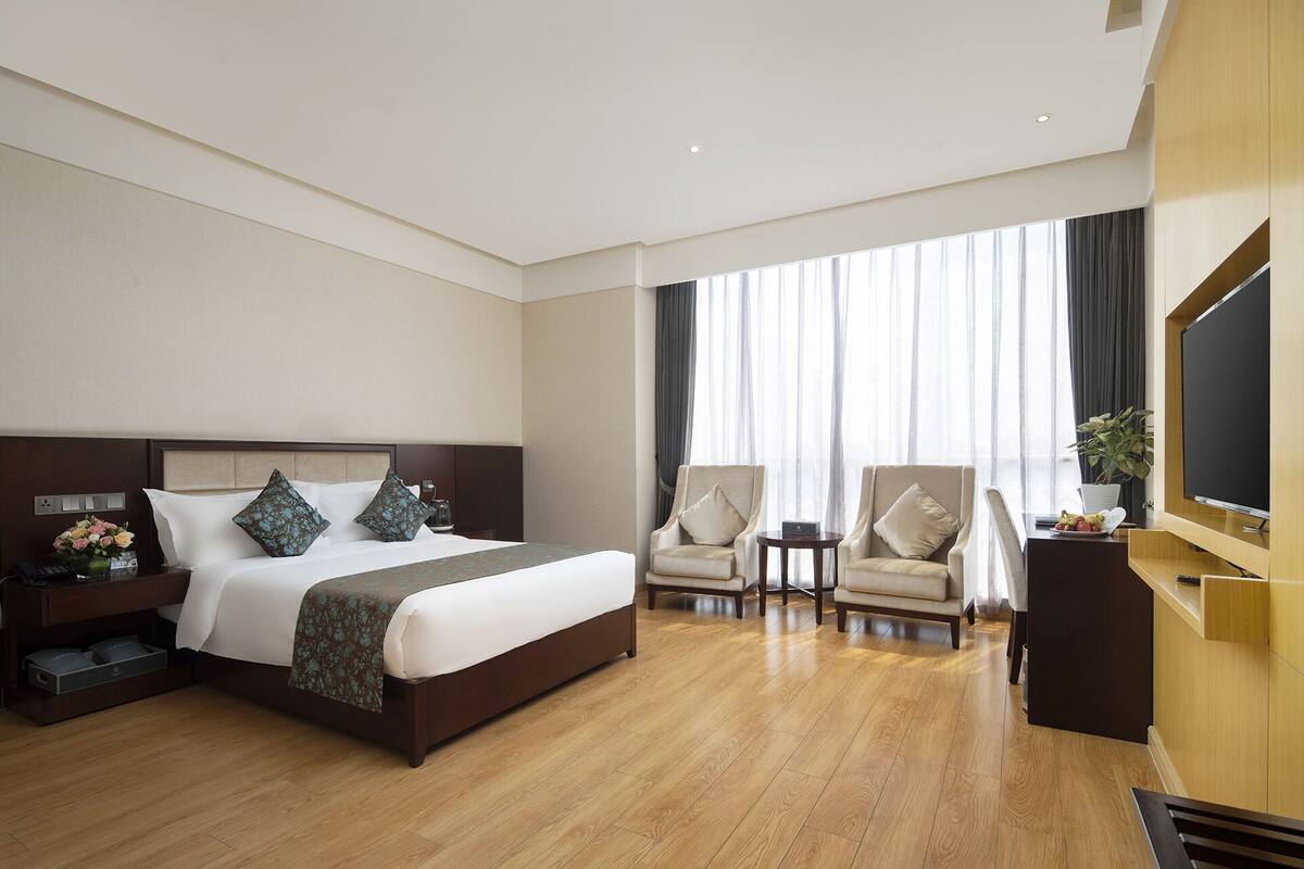 Quarto de hotel com uma cama grande, duas poltronas, televisão e uma janela ampla com cortinas.
