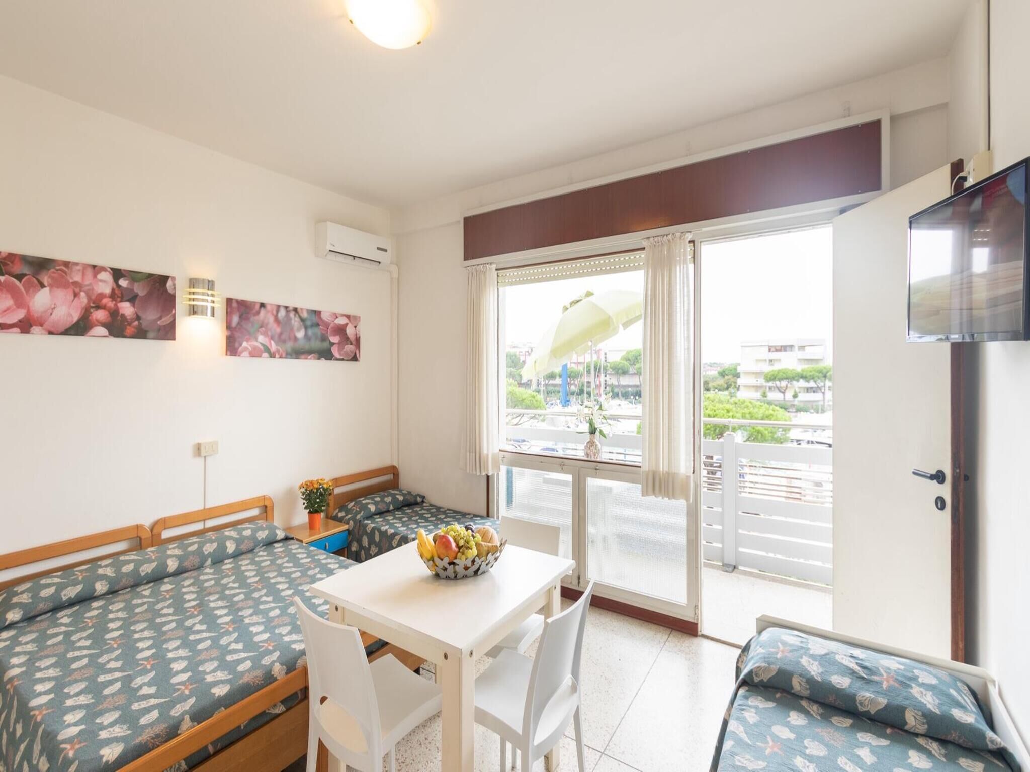Quarto compacto com cama, uma mesinha com uma fruteira, TV e varanda com vista para a cidade.
