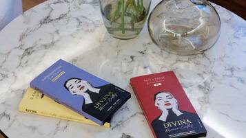 Uma mesa de mármore com um vaso de tulipas amarelas, um livro intitulado 'Divina' e um livro vermelho em um sofá branco ao fundo.