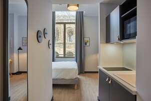 Quarto moderno com cama, janela com cortinas, área de cozinha com micro-ondas e porta que dá para outro espaço.