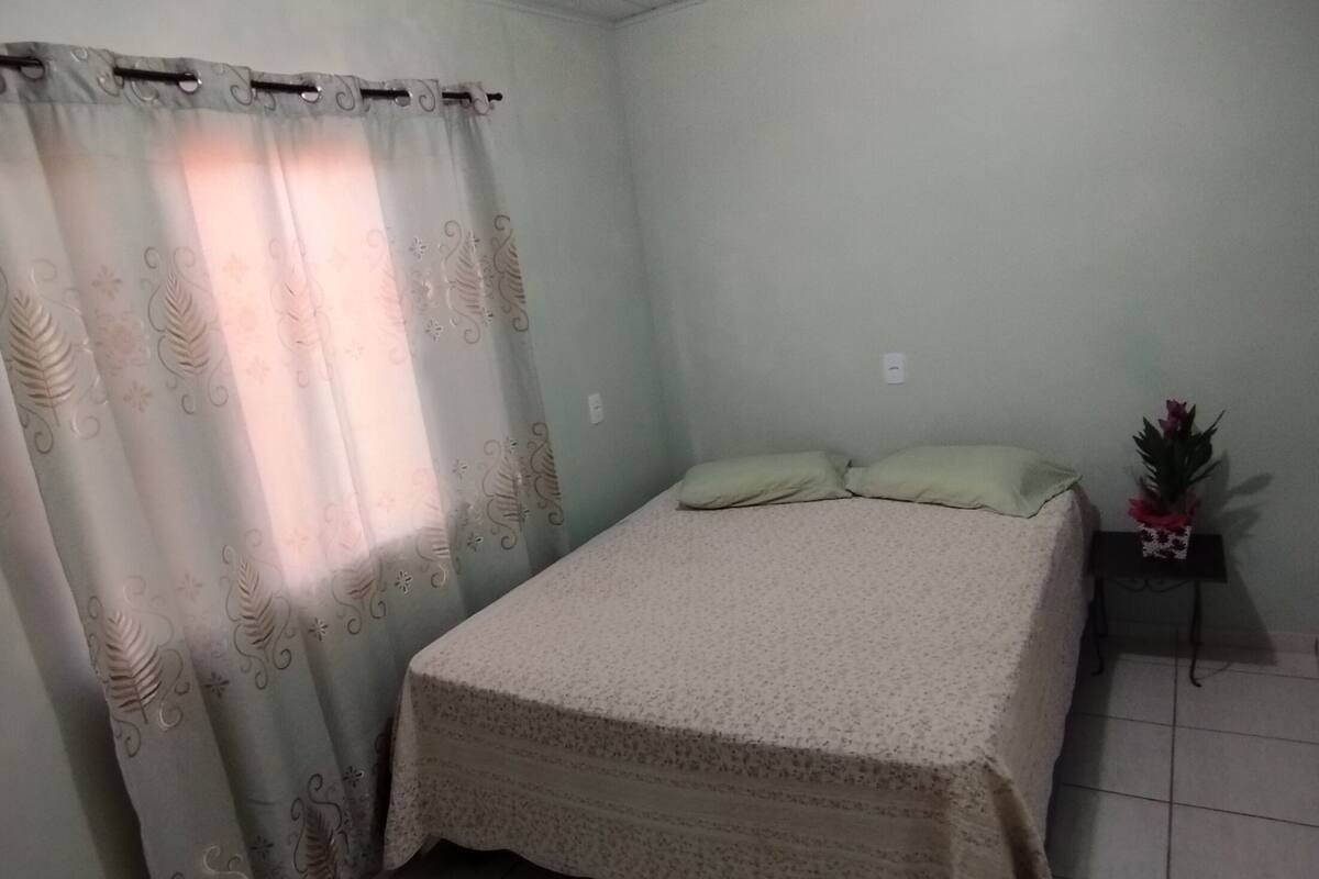 Quarto com cama, cortina e uma pequena mesa com uma flor.