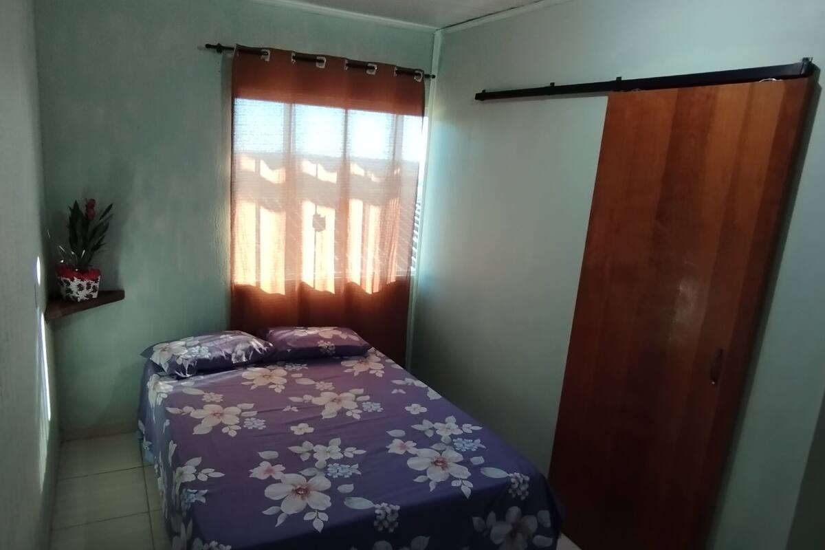 Quarto pequeno com cama, janela com cortinas, porta de madeira e prateleira na parede com uma planta em vaso.