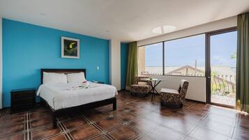 Quarto com cama, uma mesinha, duas cadeiras, mesa de cabeceira e uma janela grande com vista para edifícios.