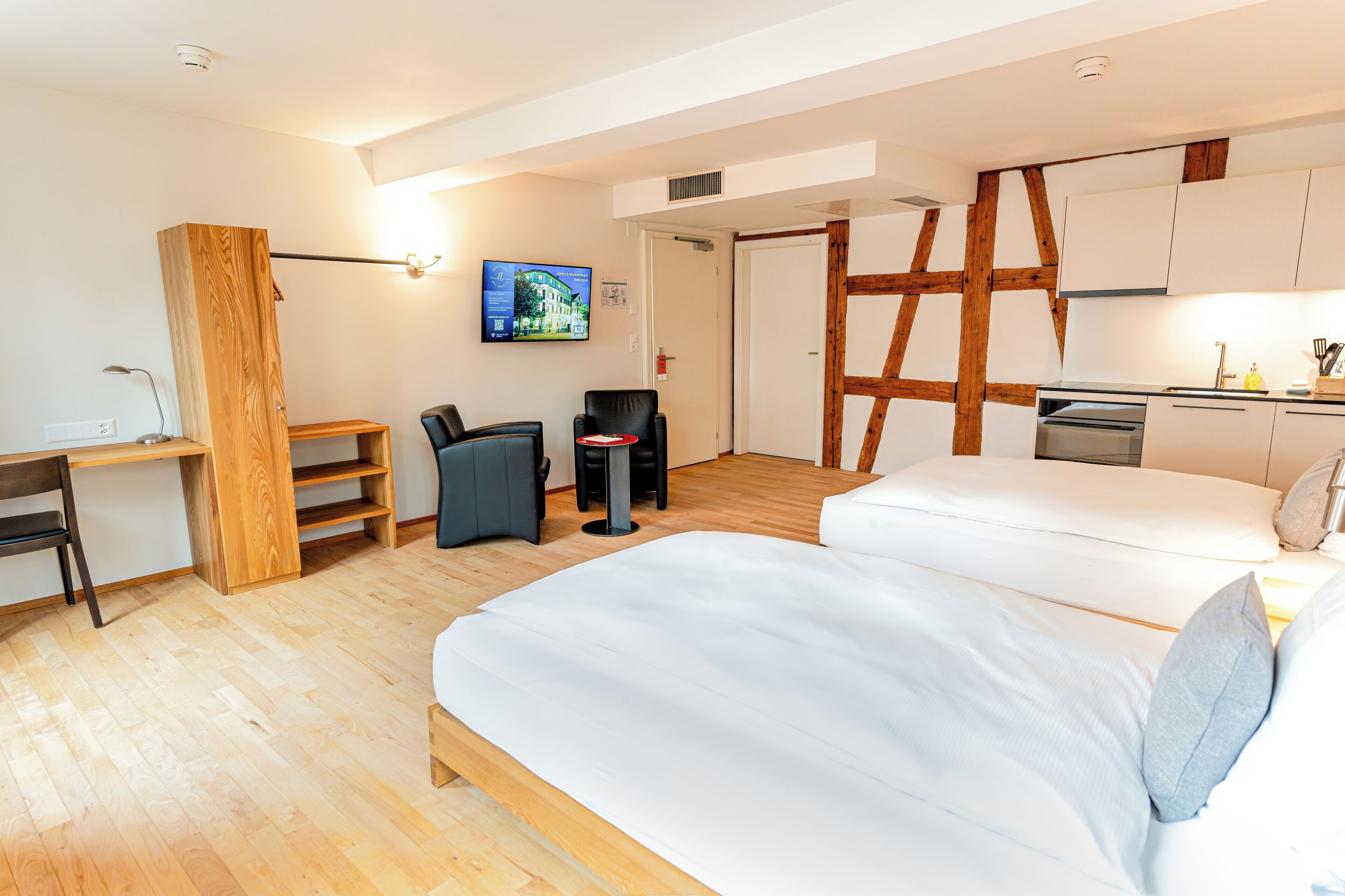 Quarto de hotel moderno com piso de madeira, cama, escrivaninha, cadeira, TV e cozinha compacta.