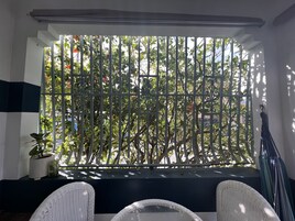 Quarto com uma janela grande, cadeiras de vime branco, uma mesinha e um vaso de planta.