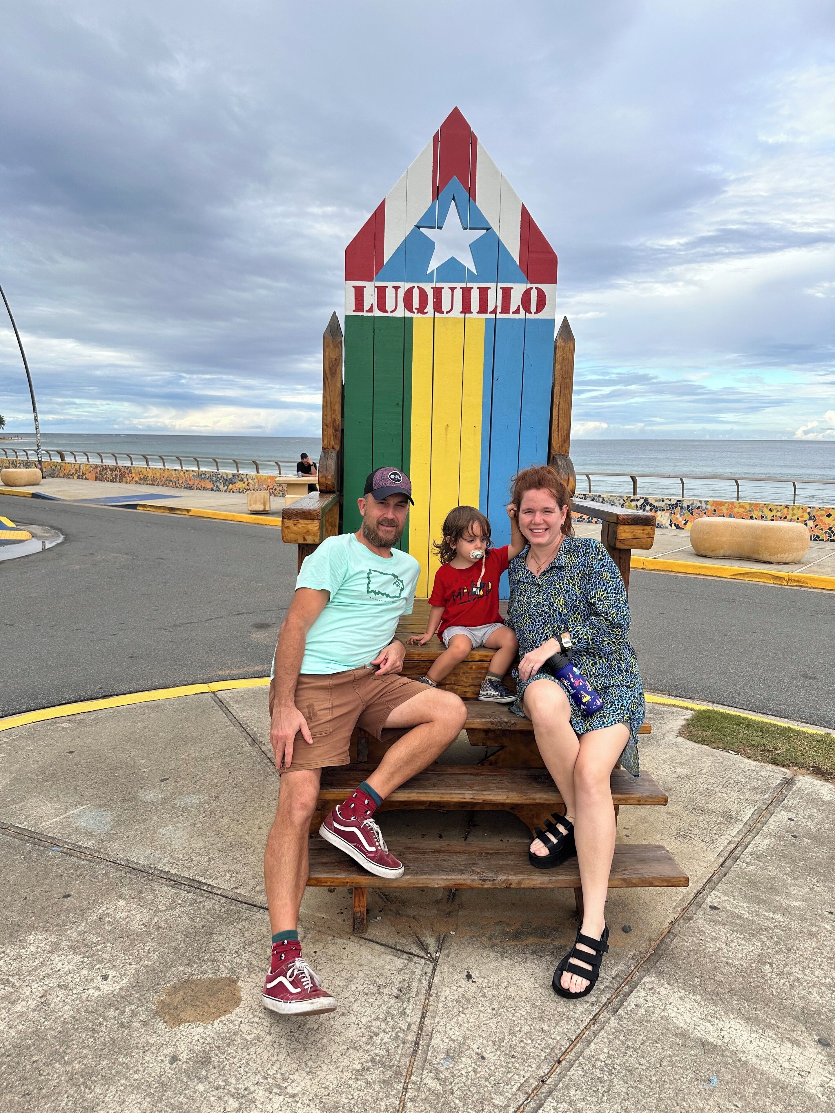 Uma família sentada num banco com uma placa colorida que diz "LUQUILLO" ao fundo.