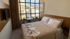Quarto de hotel com cama, televisão, janela com vista para prédios e uma pequena mesa com cadeiras.