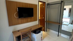 Quarto compacto com televisão montada, pequena escrivaninha de madeira, mini-geladeira e prateleira retrátil.