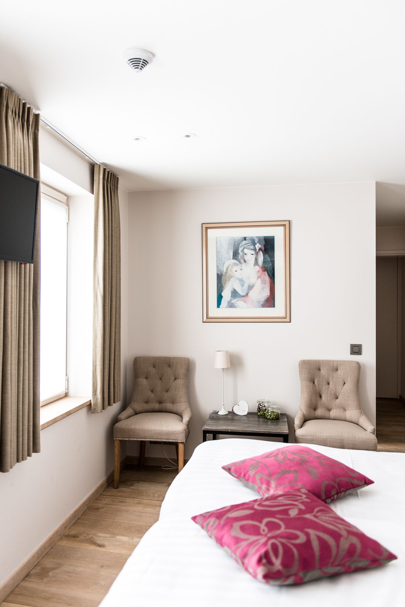 Quarto de hotel com uma cama, duas poltronas, uma mesinha de centro com abajur, um quadro na parede e uma televisão fixada na parede.