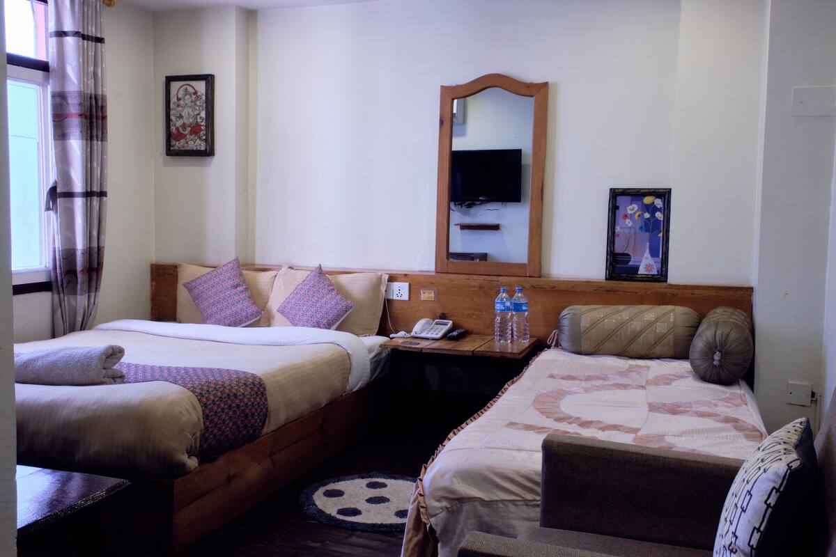 Quarto de hotel com duas camas, cabeceira de madeira, espelho, mesa de cabeceira com telefone e garrafas de água, e tapete redondo pequeno.