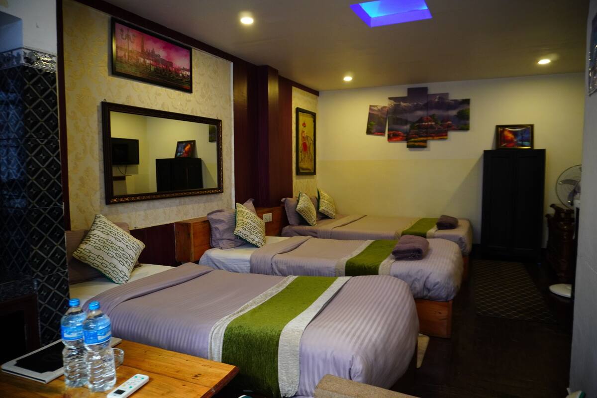 Quarto de hotel com três camas, uma mesa com um laptop e garrafas de água, um espelho e obras de arte nas paredes.