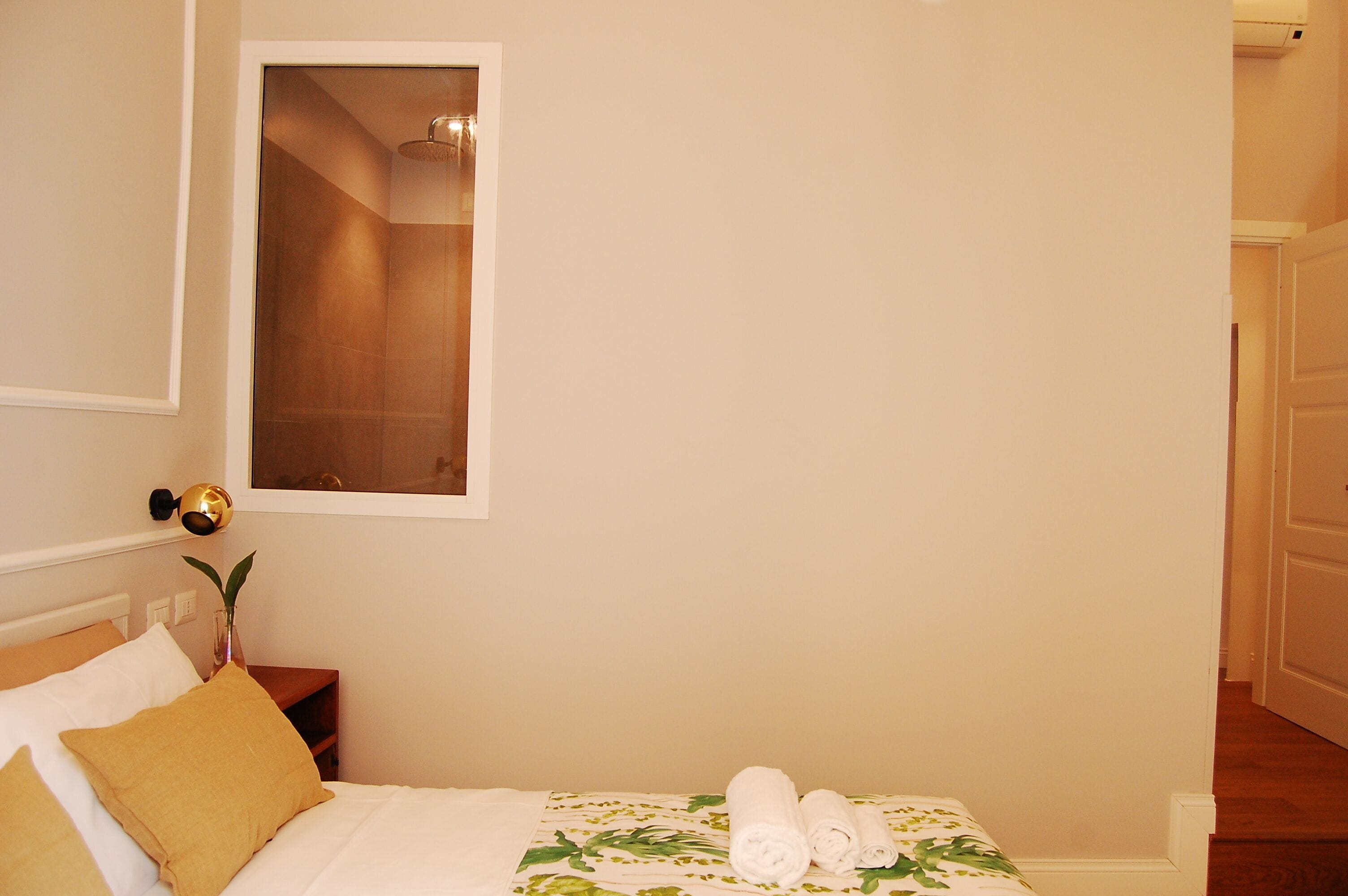 Quarto de hotel com cama, mesa de cabeceira com abajur, espelho e banheiro visível por uma porta aberta.