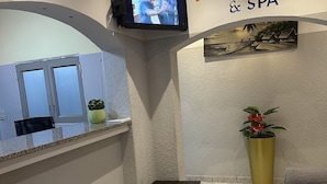 Uma área de recepção com balcão, televisão, placa de recepção, planta em vaso e uma cama de spa.