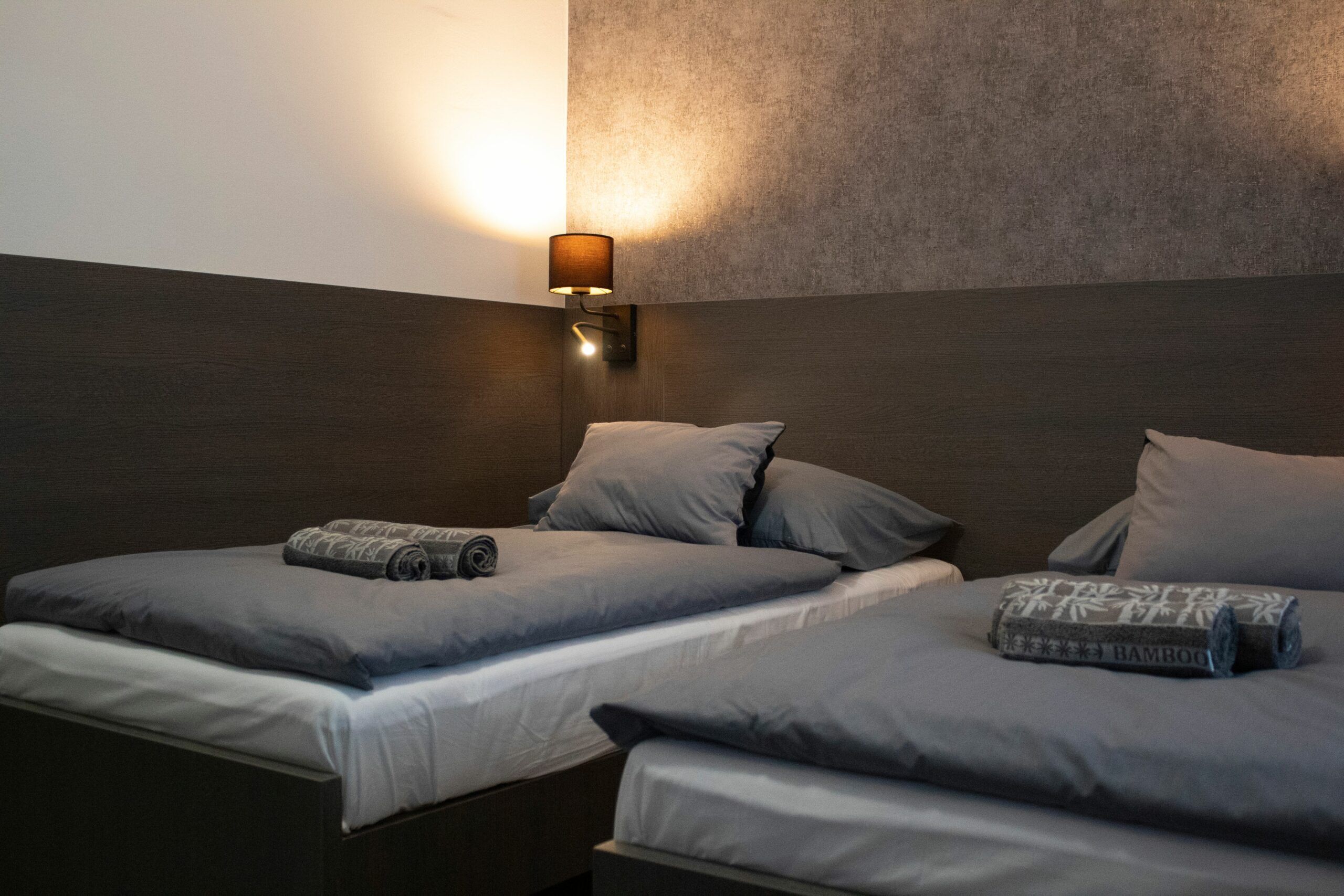 Quarto de hotel com duas camas de solteiro, uma luminária na parede e parede texturizada.