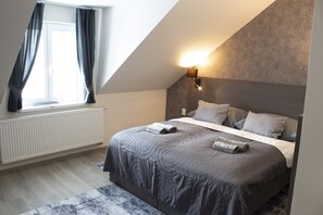 Um quarto moderno com teto inclinado, uma cama grande com colcha cinza, uma mesa de cabeceira e uma luminária fixada na parede.