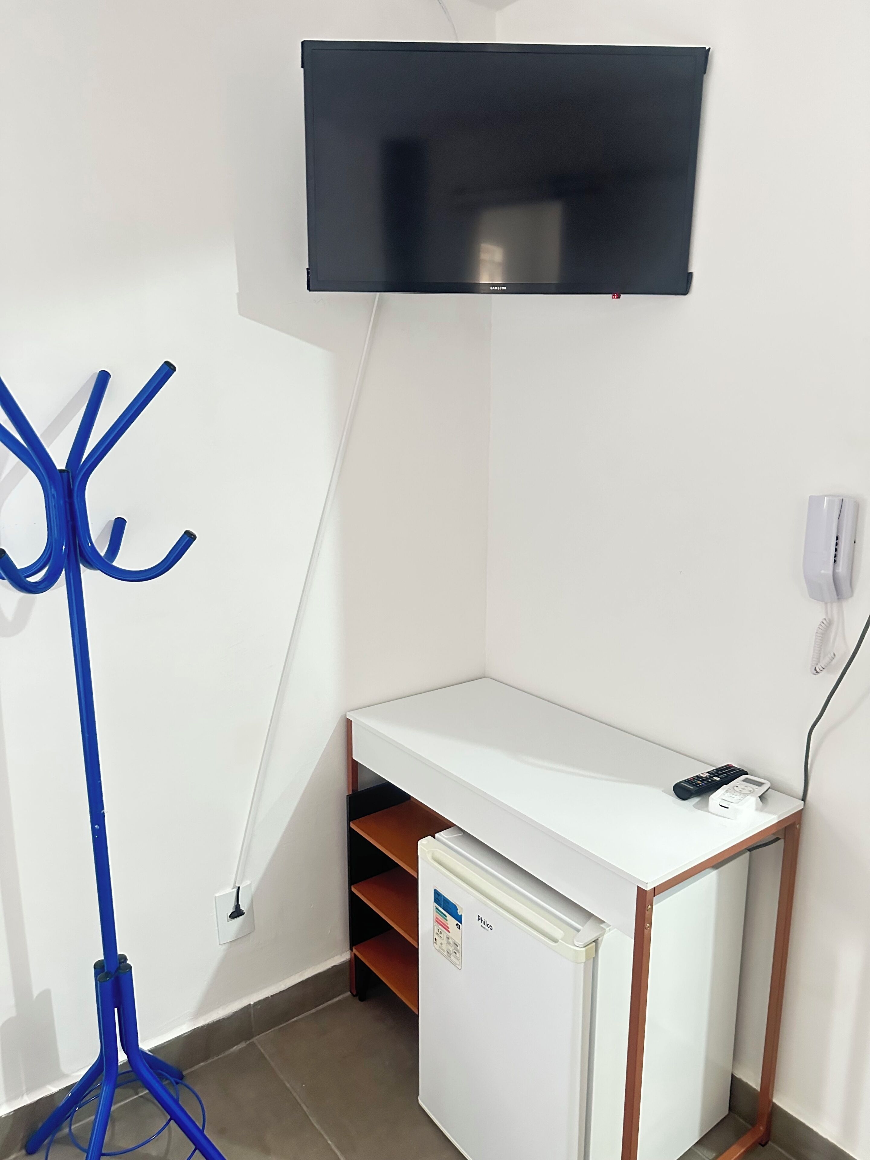 Quarto com televisão de tela plana na parede, escrivaninha branca com mini-refrigerador, abajur azul e telefone na parede.
