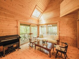 Interior acolhedor de uma cabana, com piano, área de estar e grandes janelas.