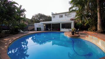 Uma piscina com um terraço coberto, rodeada por vegetação exuberante e um edifício branco com telhado plano.