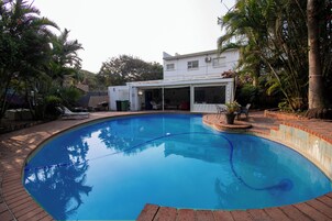 Uma piscina com um terraço coberto, rodeada por vegetação exuberante e um edifício branco com telhado plano.