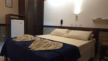 Quarto de hotel com cama, guarda-roupa, pequena geladeira, mesa de cabeceira e telefone fixado na parede.