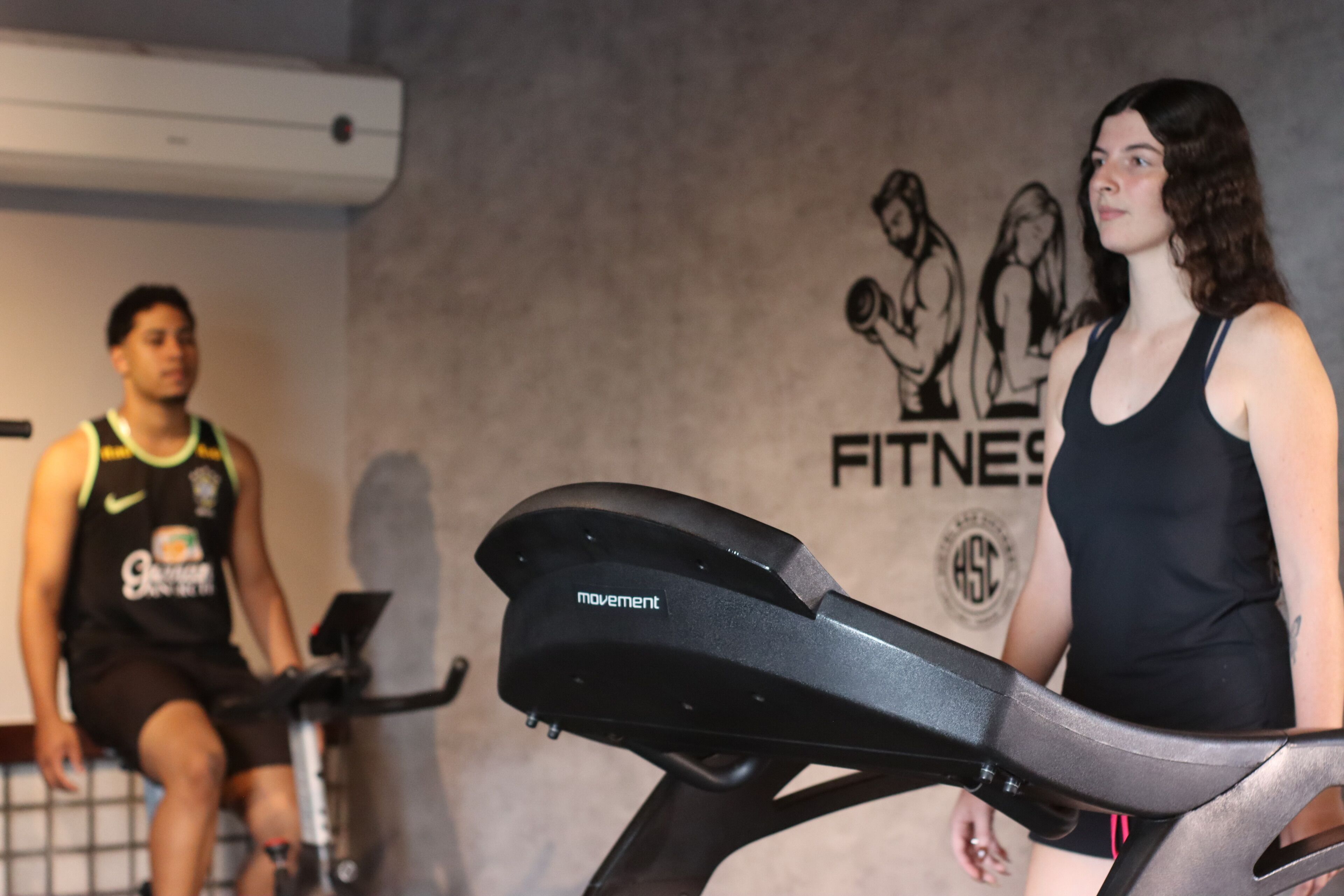 Uma academia com bicicletas ergométricas e uma parede com ilustrações relacionadas a exercícios físicos.