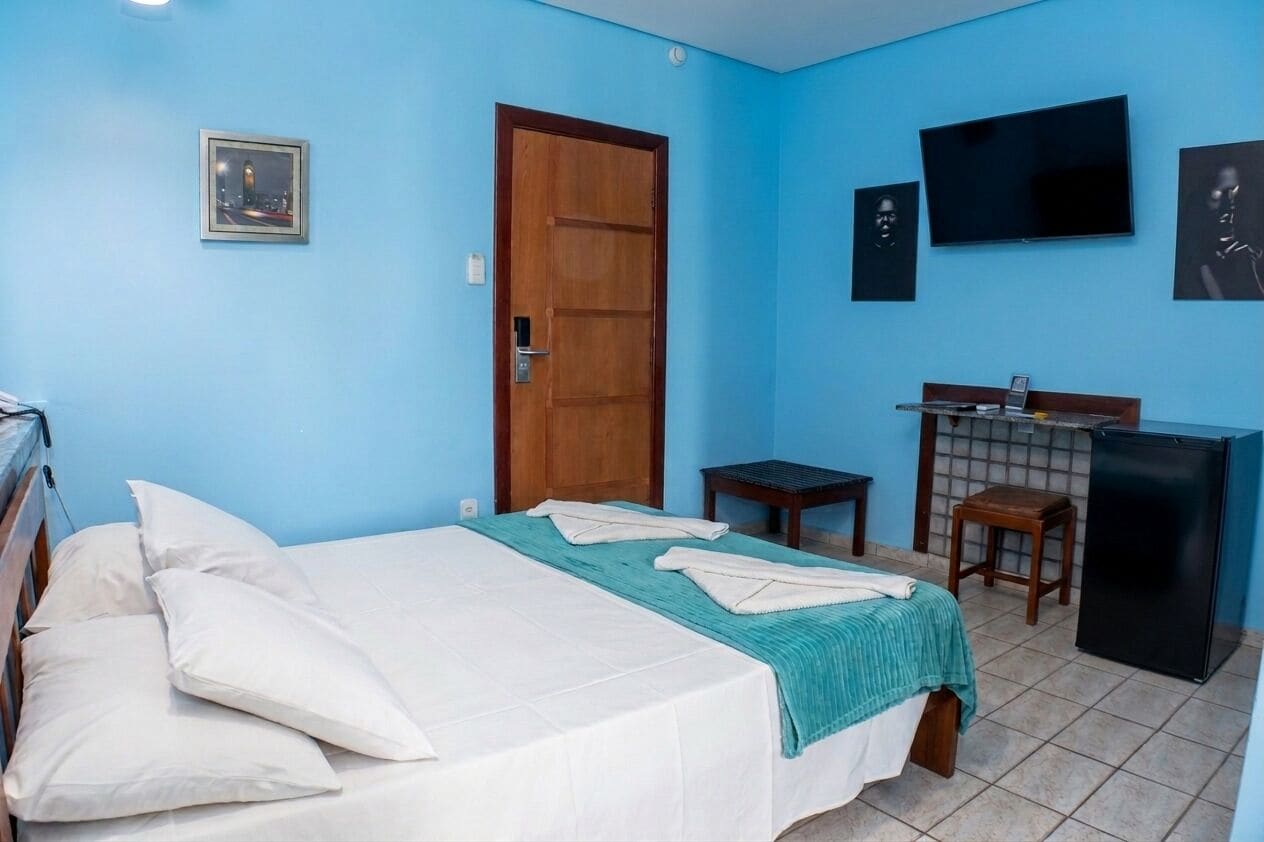 Quarto com uma cama, uma escrivaninha, uma televisão e uma porta.