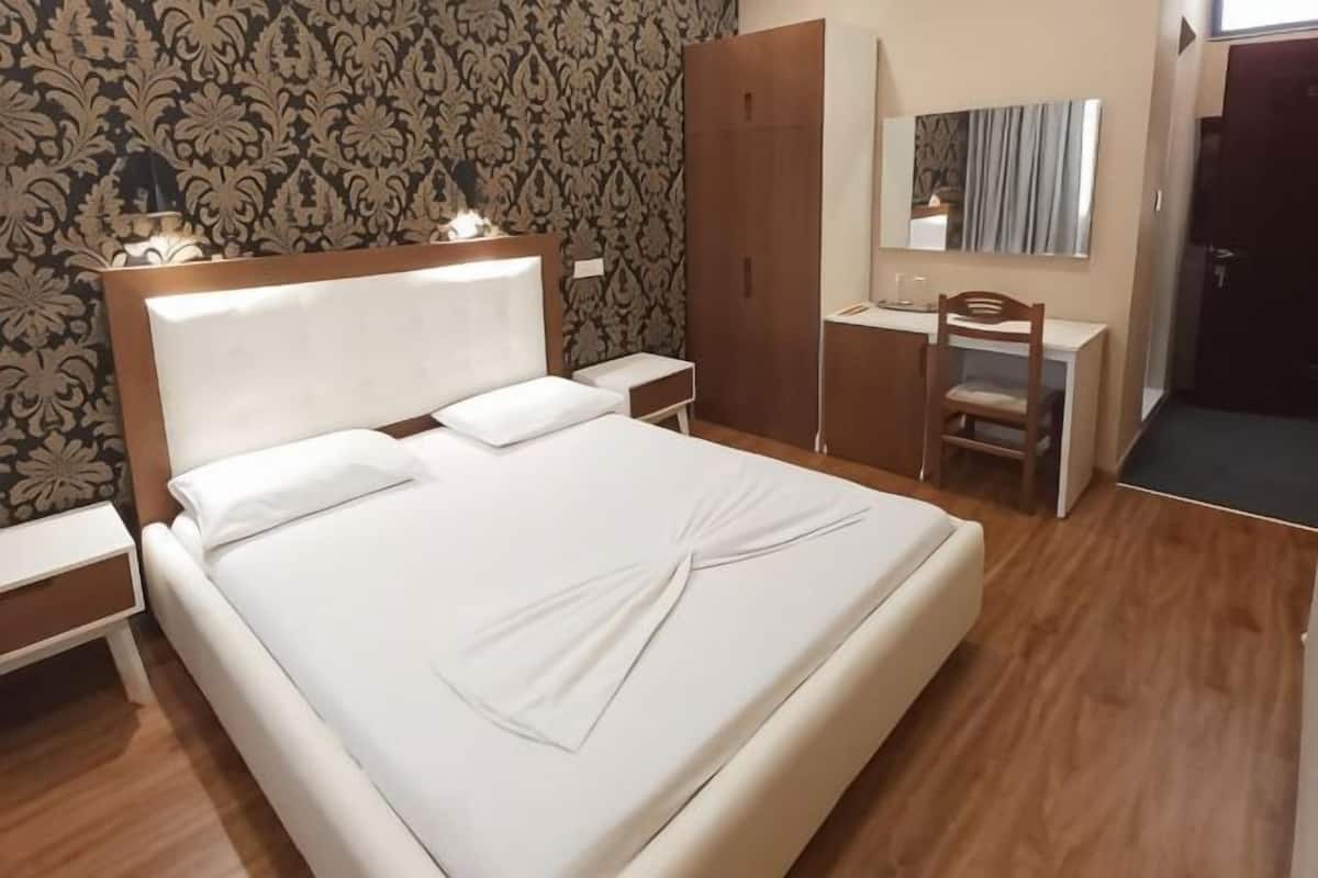 Quarto de hotel com cama, mesas de cabeceira, escrivaninha, cadeira, guarda-roupa e espelho.