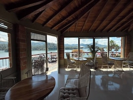 Amplo espaço interno com pé direito alto, grandes janelas com vista para um lago e montanhas, e diversos arranjos de assentos incluindo mesas e cadeiras.
