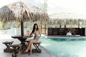 Área da piscina com mesa de madeira e guarda-sol de palha, zona de bar com bartender e vista para a água.