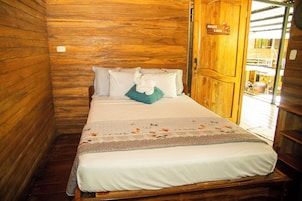 Quarto pequeno e acolhedor com paredes de madeira, cama com colchão branco e manta estampada com flores, um travesseiro verde e uma placa indicando 'ROOM LAPA'.