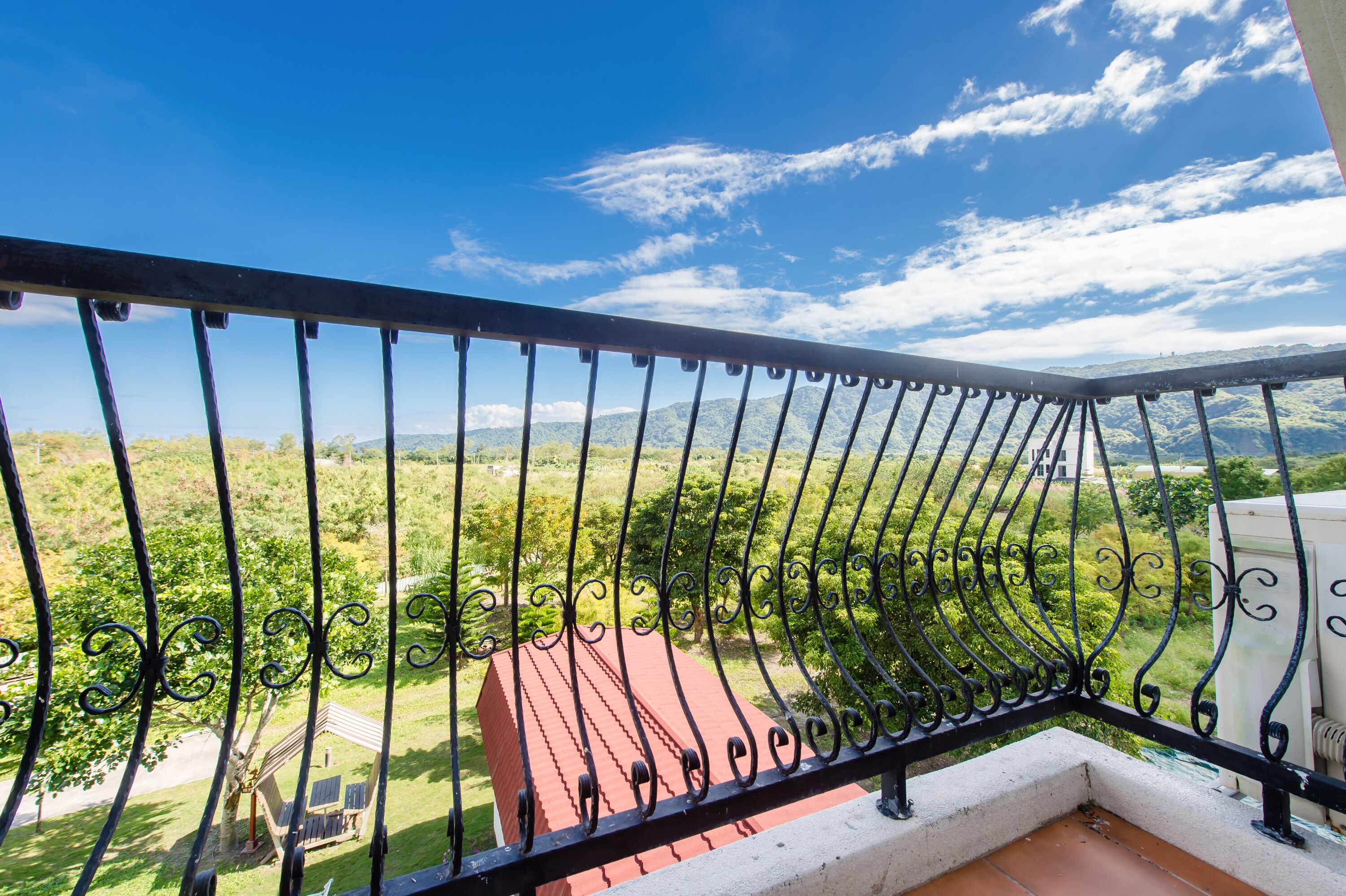 Uma varanda com grade de ferro forjado, com vista para uma paisagem verde com árvores e um edifício de telhado vermelho.