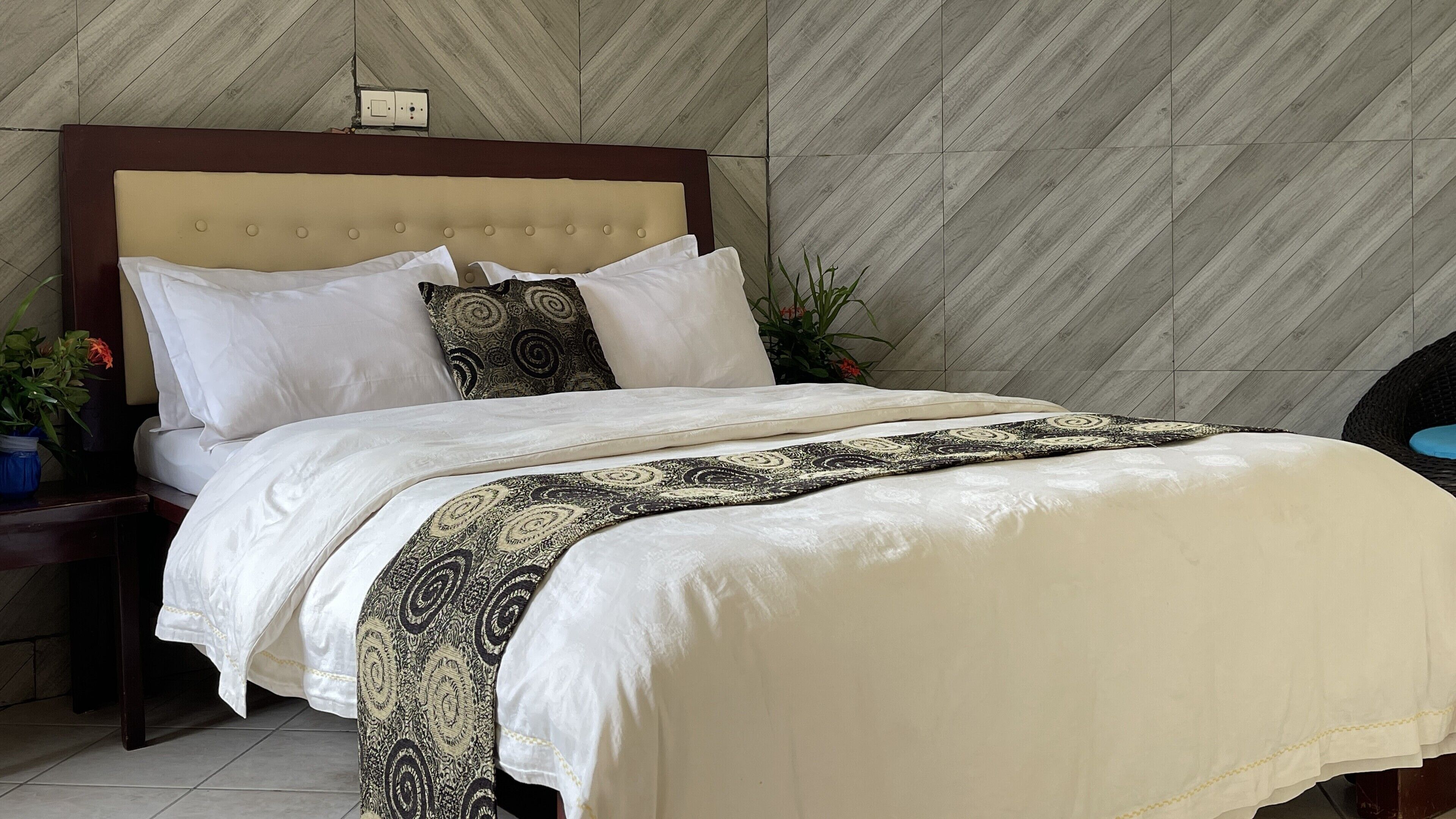 Cama bem arrumada com manta estampada, cabeceira e mesa de cabeceira com uma planta.