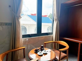 Quarto com uma mesa de madeira redonda, duas cadeiras, uma janela com cortinas e vista para edifícios com telhados azuis.