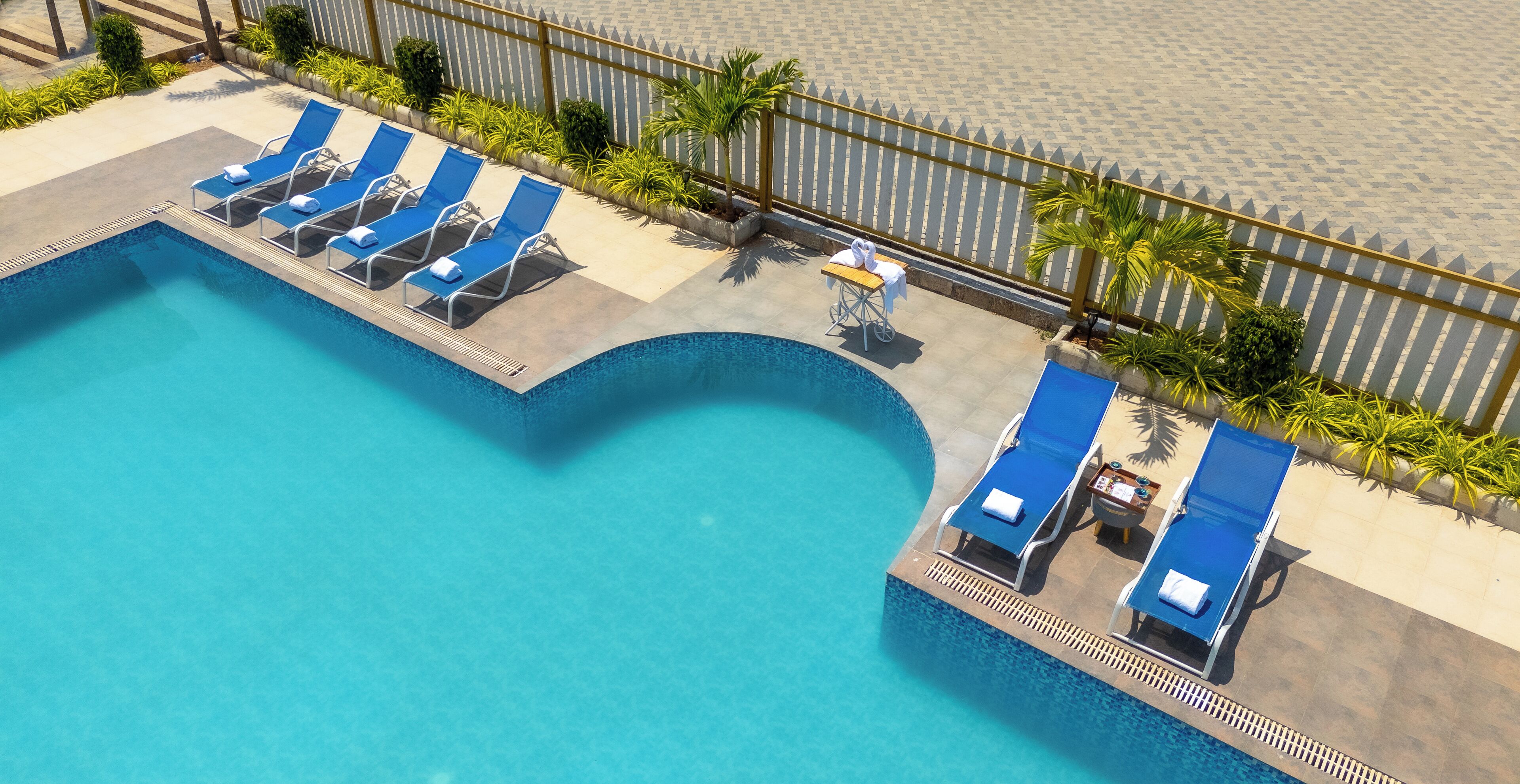 Uma piscina com cadeiras de descanso azuis e espreguiçadeira, cercada por uma grade e palmeiras.