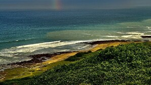 Paisagem costeira com um arco-íris sobre o oceano, uma praia com vegetação e um céu nublado.