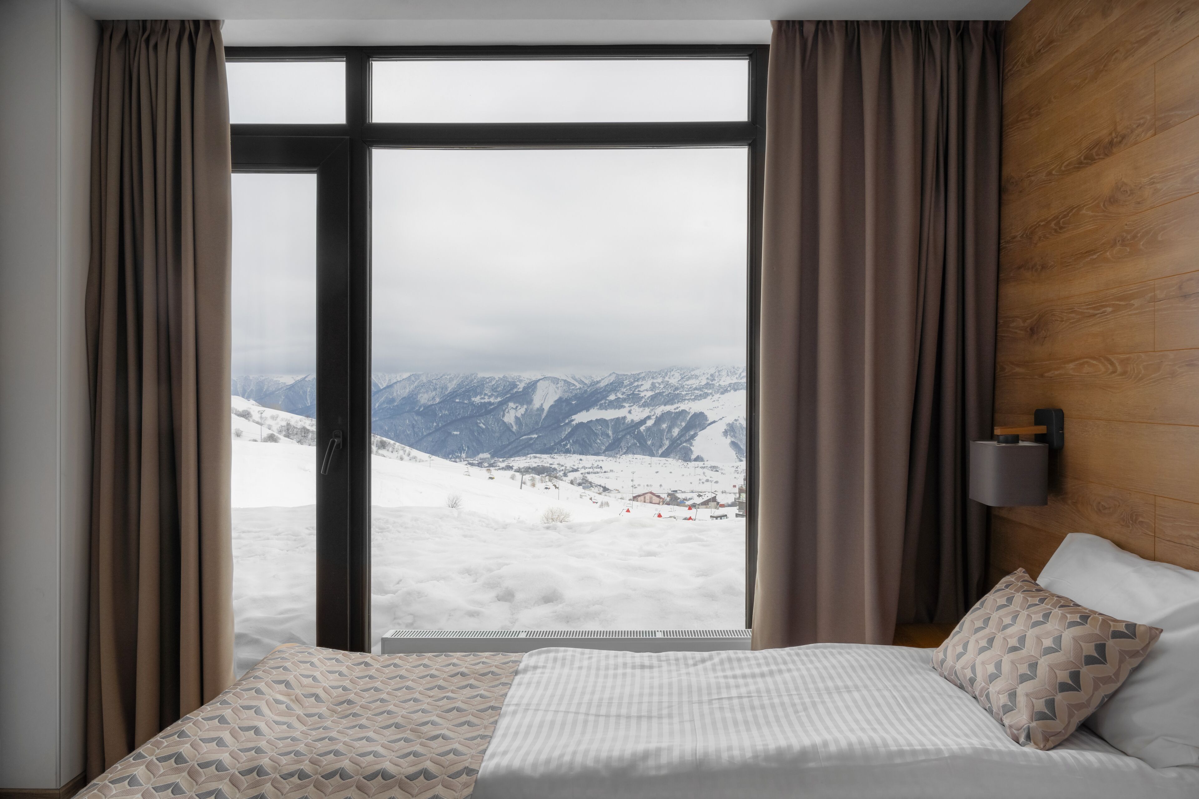 Quarto de hotel com uma janela grande que oferece vista para as montanhas, uma cama com roupa de cama estampada e um painel de parede de madeira.