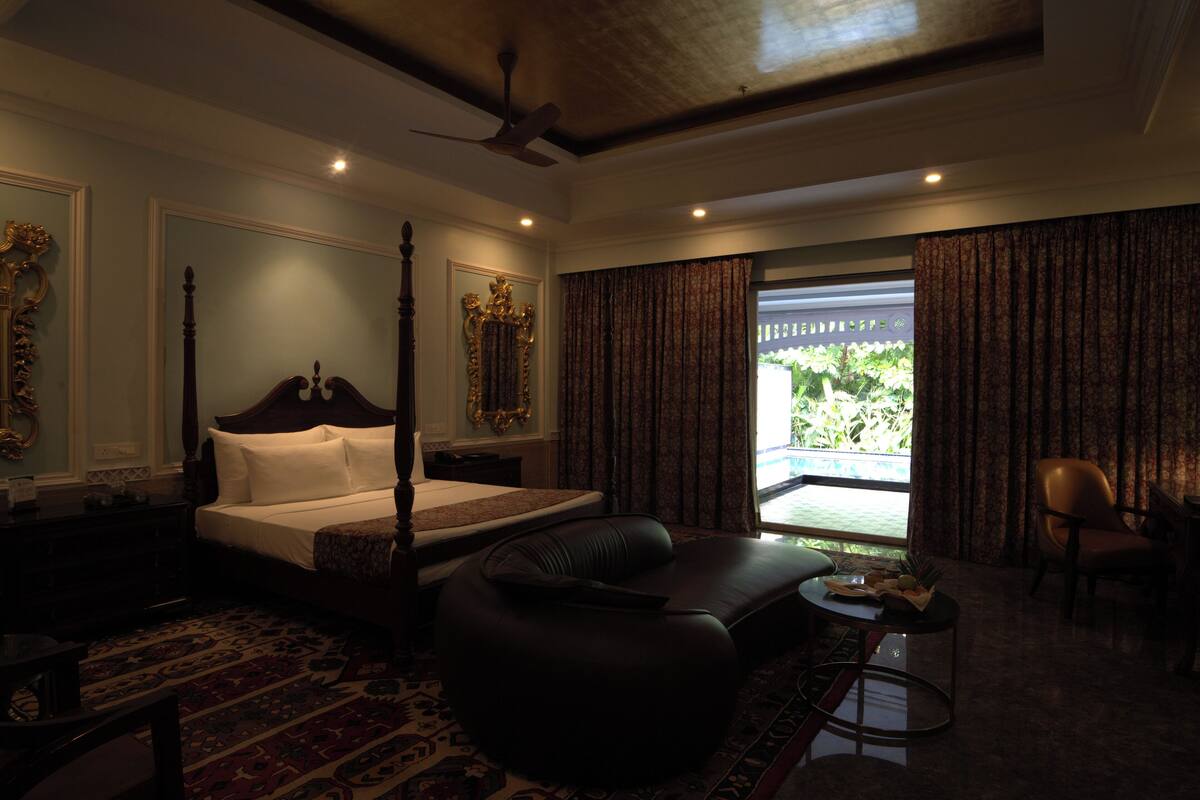 Um quarto espaçoso com uma cama de dossel, um espaço de estar com um sofá e vista para um jardim exuberante através de grandes janelas.