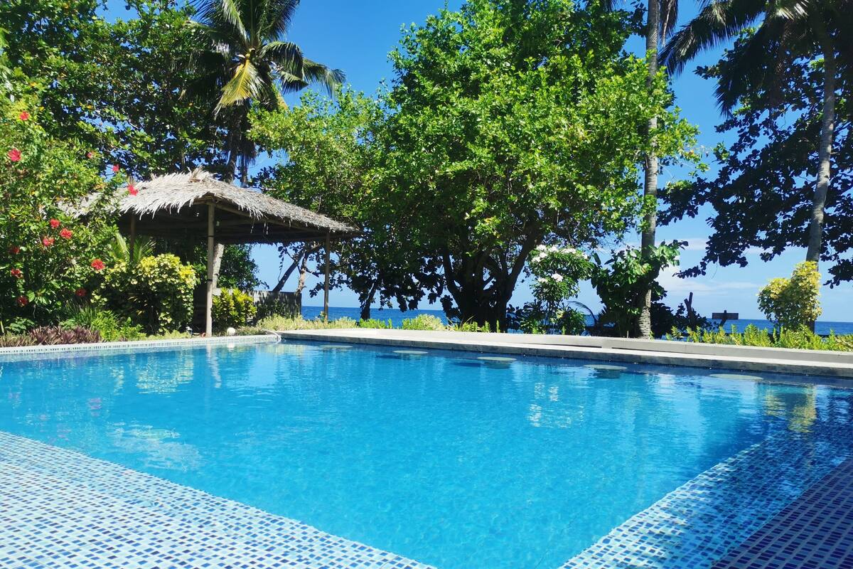 Uma piscina azul cristalina rodeada por vegetação exuberante e uma construção com telhado de palha.