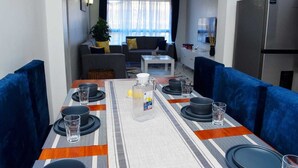 Uma sala de jantar com uma mesa posta para quatro pessoas, um sofá ao fundo e uma área de cozinha com geladeira.