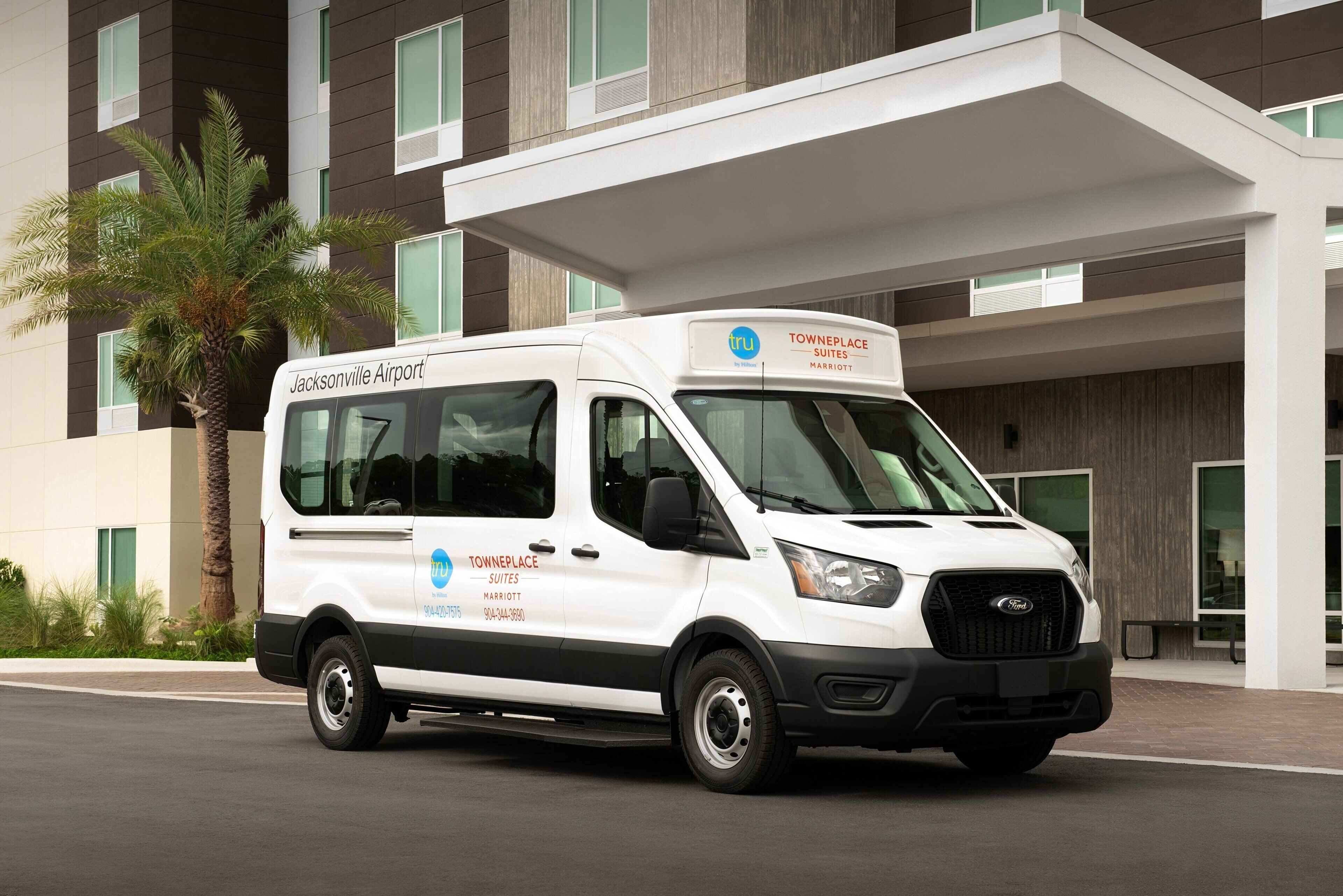 Uma van branca com a inscrição "TOWNEPLACE SUITES MARRIOTT" e "Jacksonville Airport", estacionada em frente a um moderno edifício de hotel.