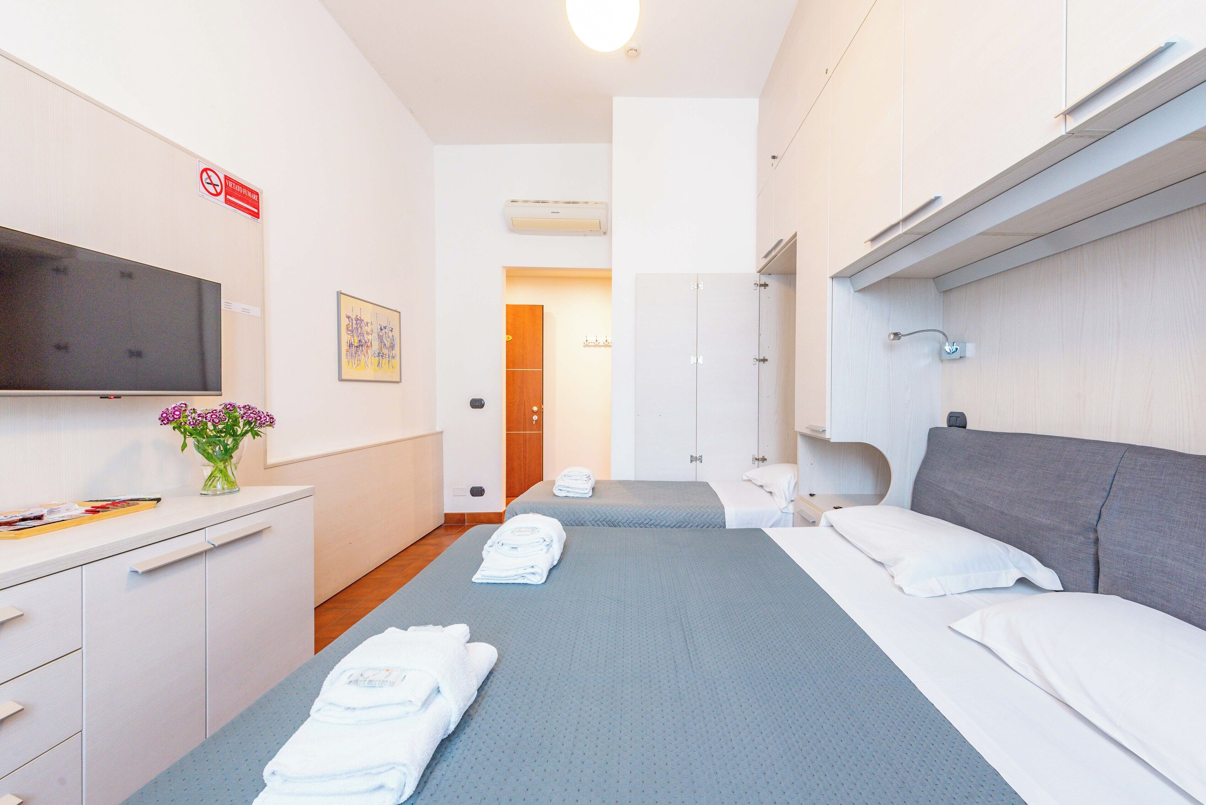 Quarto de hotel com cama, televisão, escrivaninha com um vaso de flores e banheiro visível através de uma porta aberta.