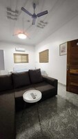 Uma sala de estar com um sofá marrom escuro, uma mesa de centro redonda pequena, um ventilador de teto e uma porta à direita.