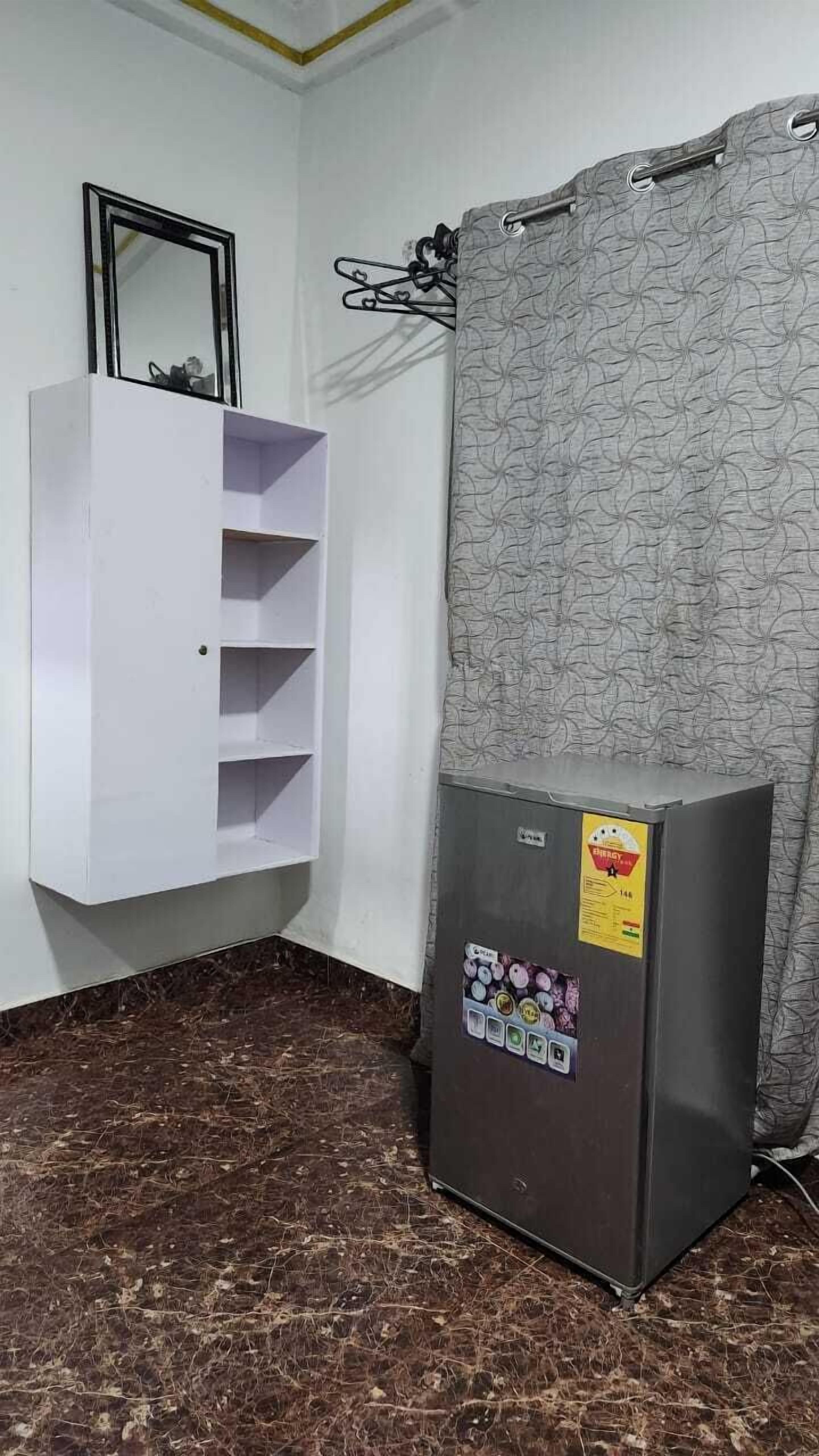 Quarto pequeno com guarda-roupa branco, cortina cinza estampada e um frigorífico pequeno com autocolantes.