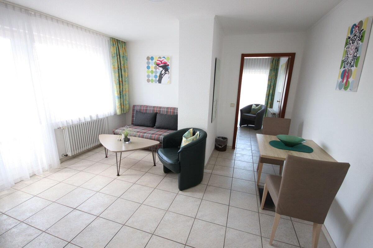 Uma sala de estar com sofá, mesa de centro, mesa de jantar com cadeiras, radiador e janela com cortinas.